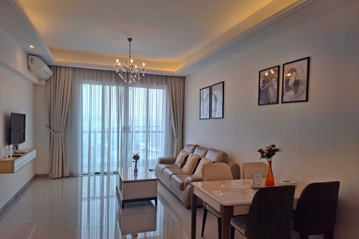 Uma sala de estar moderna com sofá, mesa de jantar e cadeiras. Há uma TV em um suporte, um lustre e uma janela grande com cortinas.