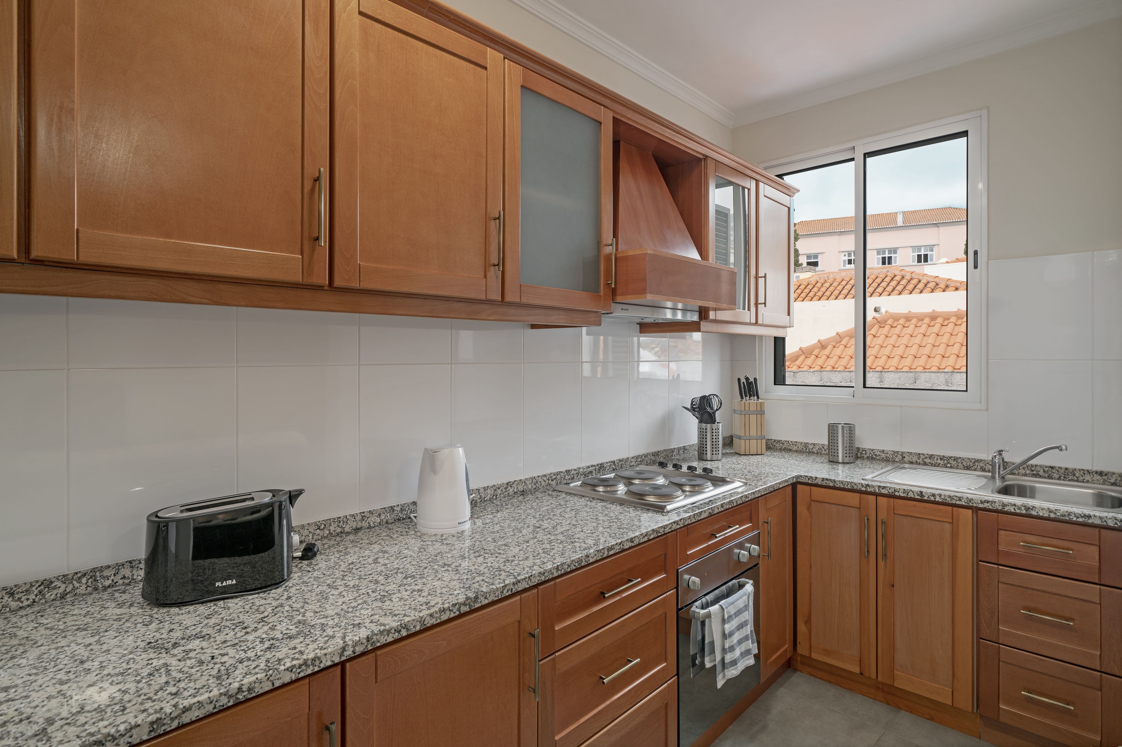 Cozinha com armários de madeira, bancada de granito, torradeira, chaleira e janela com vista para edifícios.