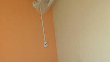Uma cama de solteiro com colchão branco, travesseiro branco, manta branca e roupão branco sobre a cama. Há um ventilador de chão branco fixado na parede acima da cama.