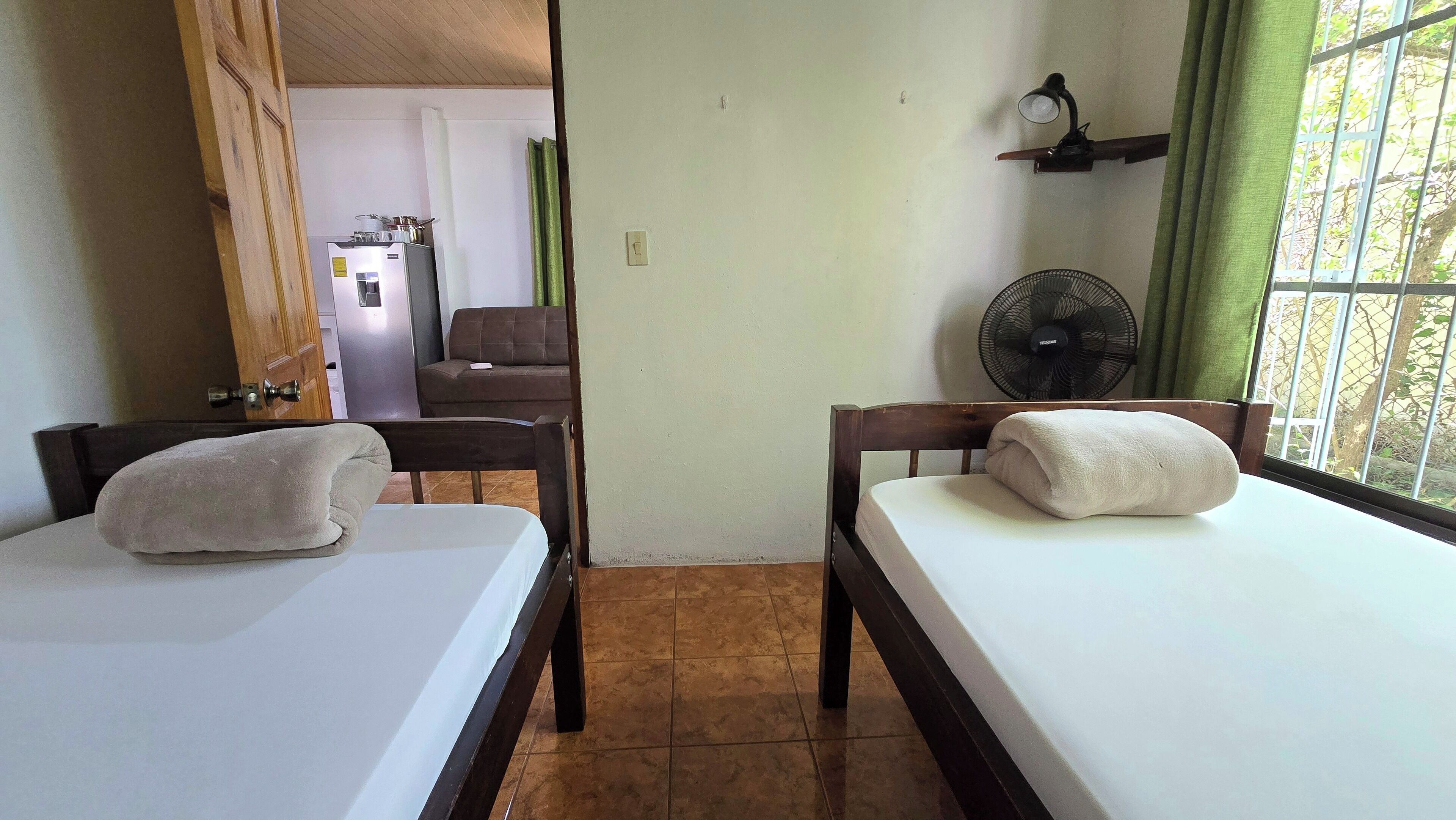 Quarto com duas camas, um ventilador e uma janela com vista para o verde.