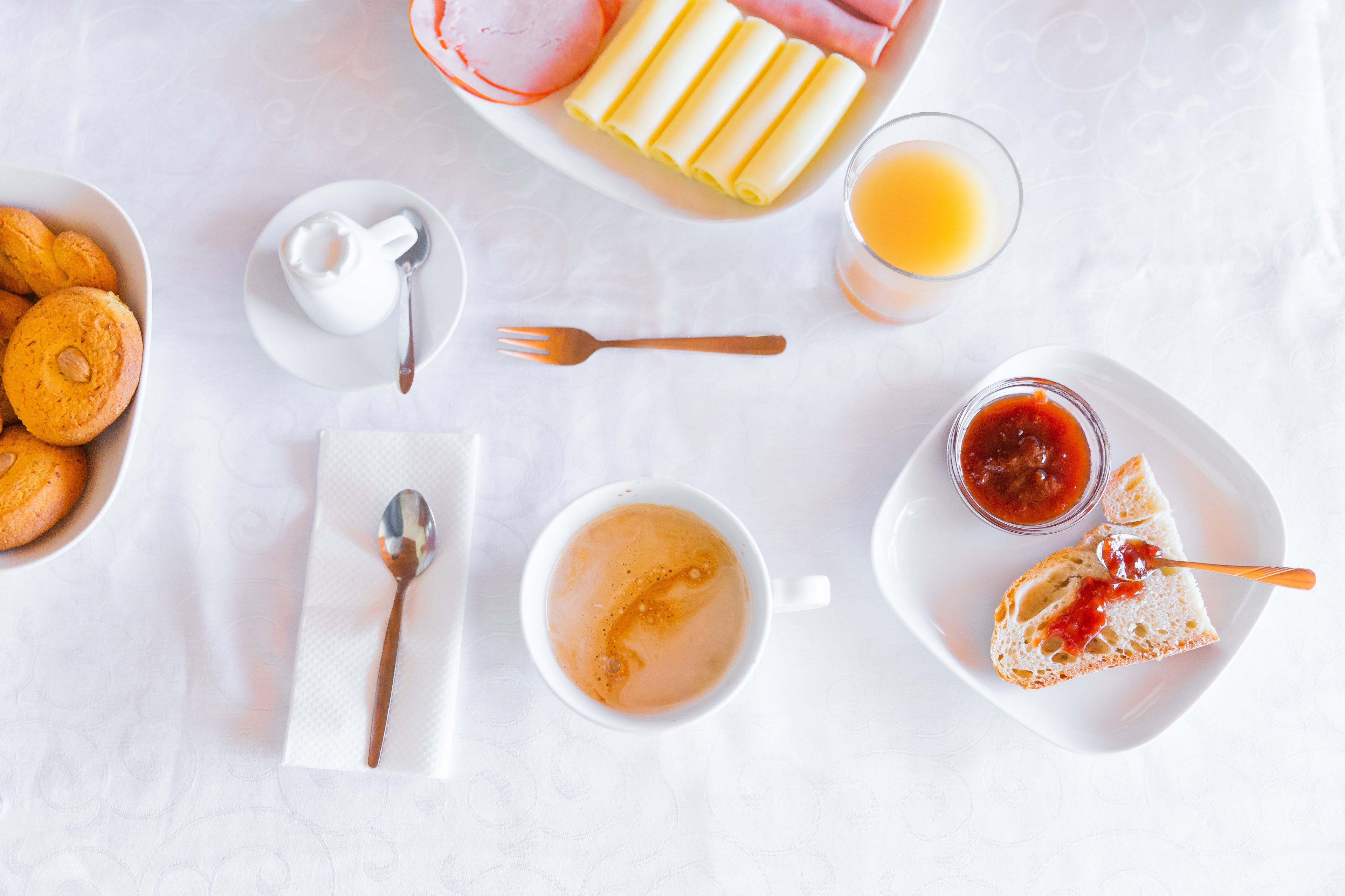 Mesa de café da manhã com panquecas, copo de suco de laranja, xícara de café, prato com geleia e torradas, e uma tigela de muffins.