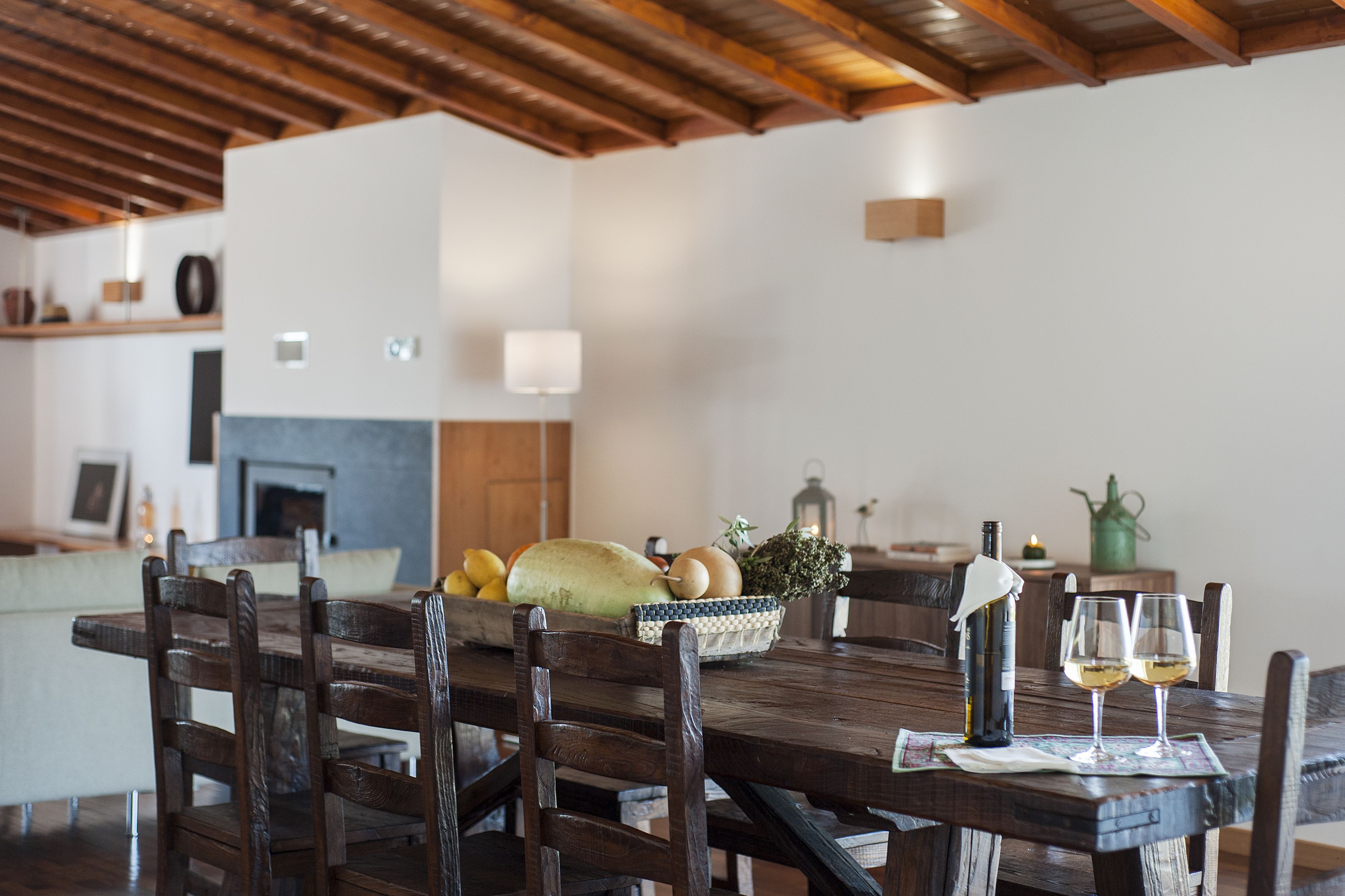 Uma mesa de jantar de madeira com cadeiras, uma cesta de frutas e dois copos de vinho branco.