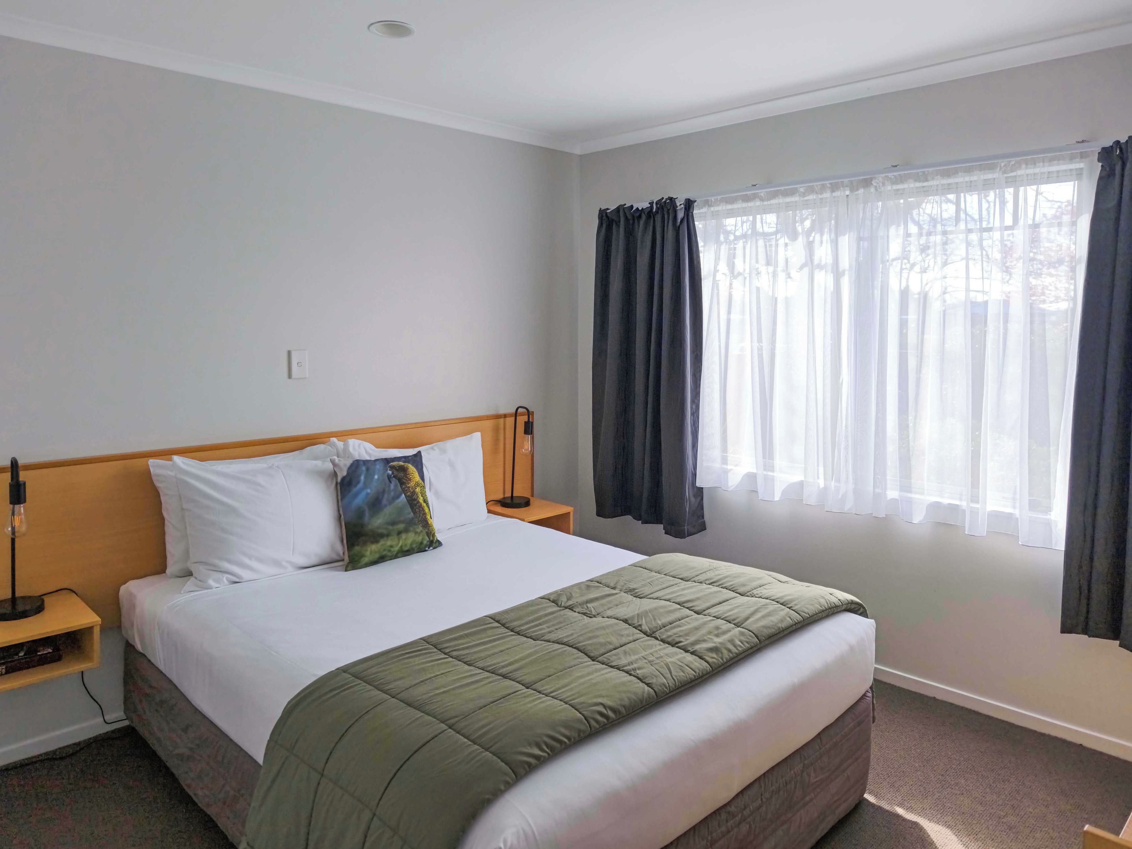 Quarto de hotel com cama, mesa de cabeceira, luminária, janela com cortinas e interruptor de luz.