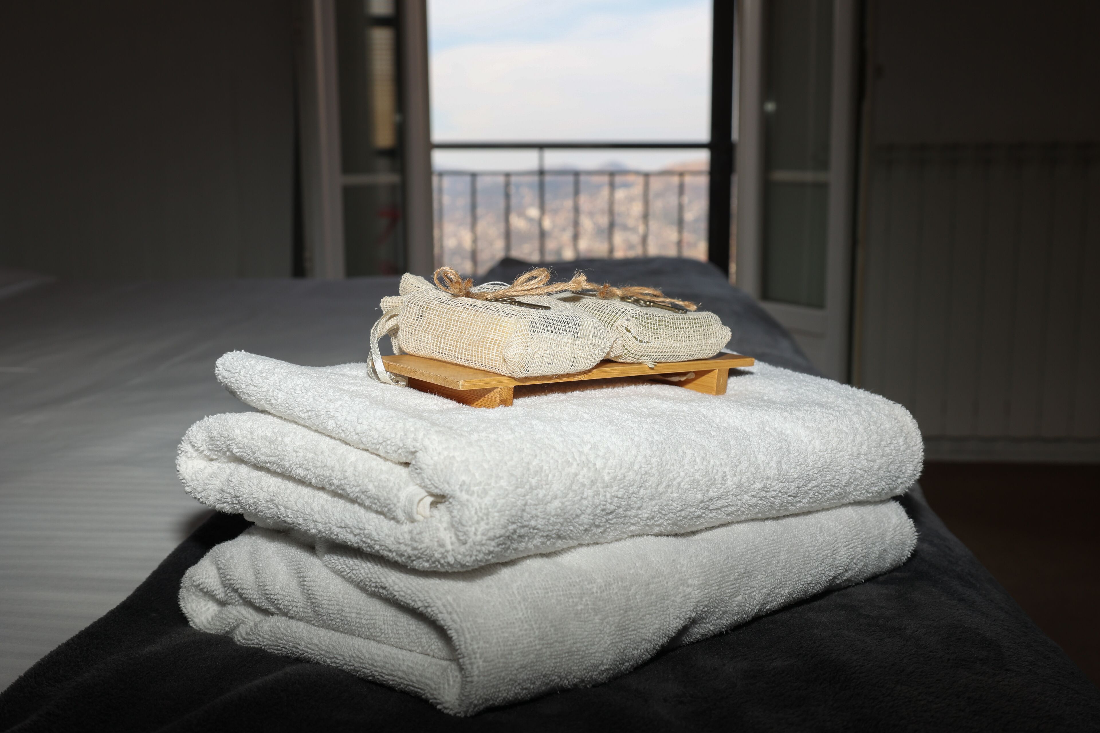 Cama com vista para uma varanda e um banquinho de madeira com uma bolsa trançada e toalhas dobradas.