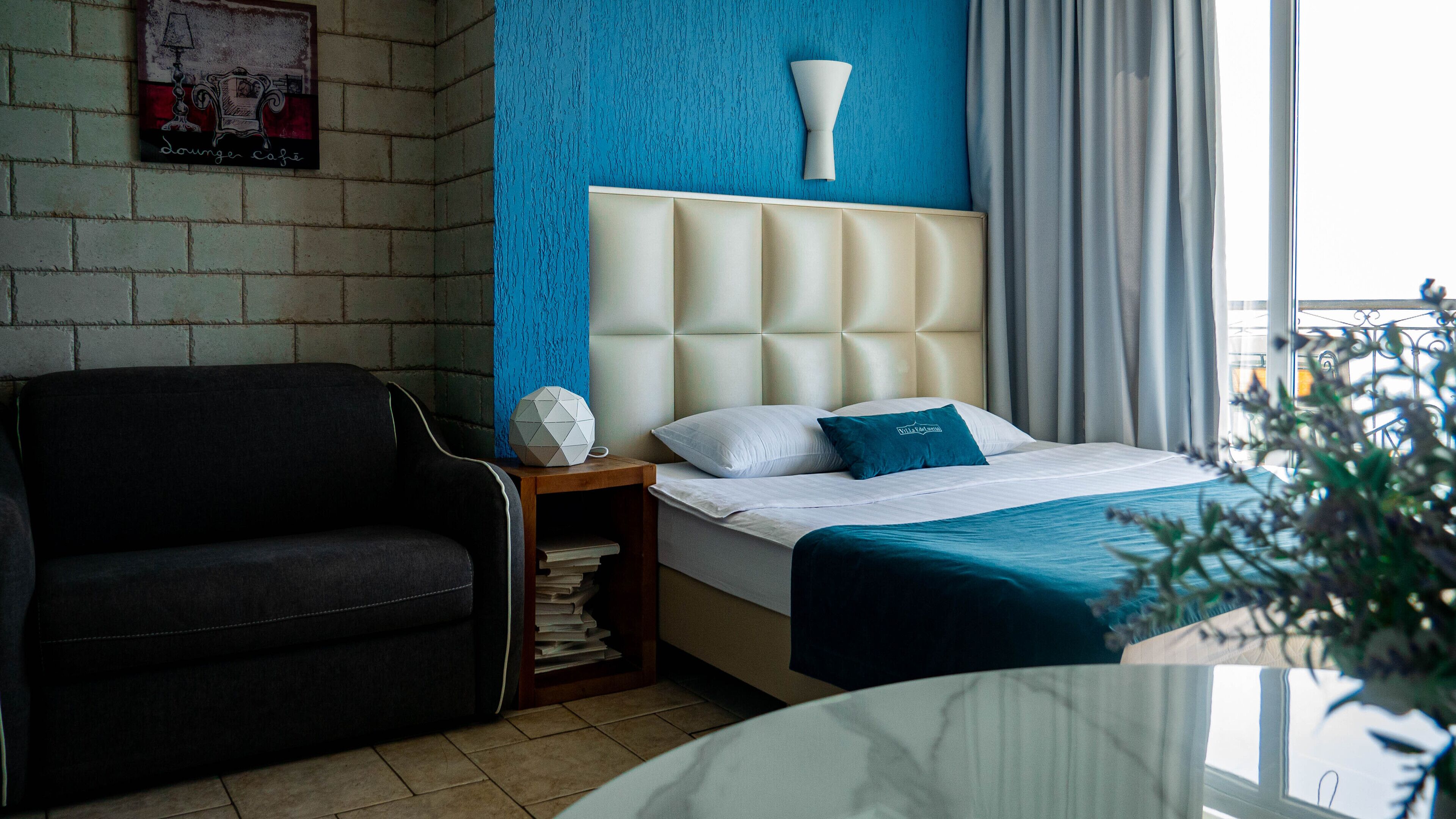 Quarto de hotel com uma cama, uma cadeira, uma mesa de cabeceira com livros, parede azul texturizada e uma janela com cortinas.
