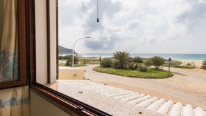 Vista da janela com vista para uma praia, área pavimentada, palmeiras e um poste de luz.