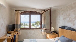 Quarto de hotel com cama, escrivaninha com televisão, janela com cortinas e vista para o mar.