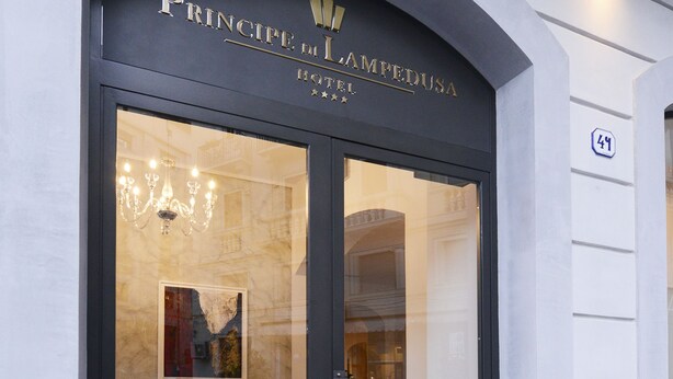 A entrada do Hotel Prince de Lampedusa, com uma porta em arco imponente, fachada de vidro refletiva e um lustre acima.