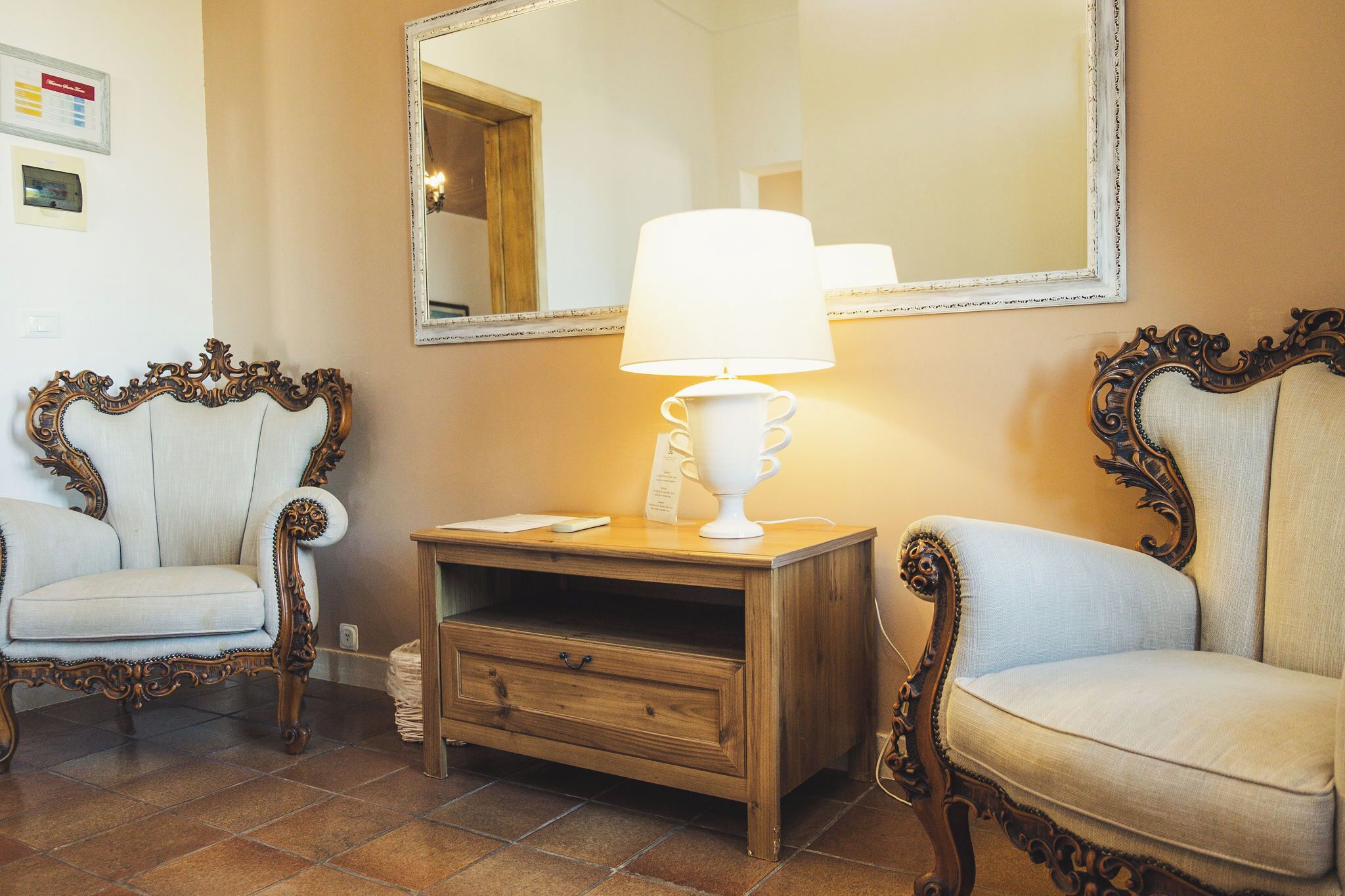 Um quarto com duas cadeiras ornamentadas, uma mesa lateral de madeira com abajur e um espelho na parede.