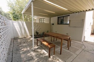 Uma área de pátio ao ar livre com mesa e bancos de madeira, cerca branca e um espaço coberto para sentar.