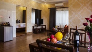 Quarto de hotel com área de jantar, televisão, minibar e uma cesta de frutas.