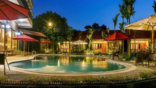 Área de piscina com espreguiçadeiras e guarda-sóis, cercada por palmeiras e um edifício com telhado em azulejos.