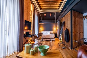 Quarto de hotel moderno com piso de madeira, uma cama grande, uma escrivaninha e uma cadeira. Há duas luminárias de cada lado da cama e um cabideiro na parede.