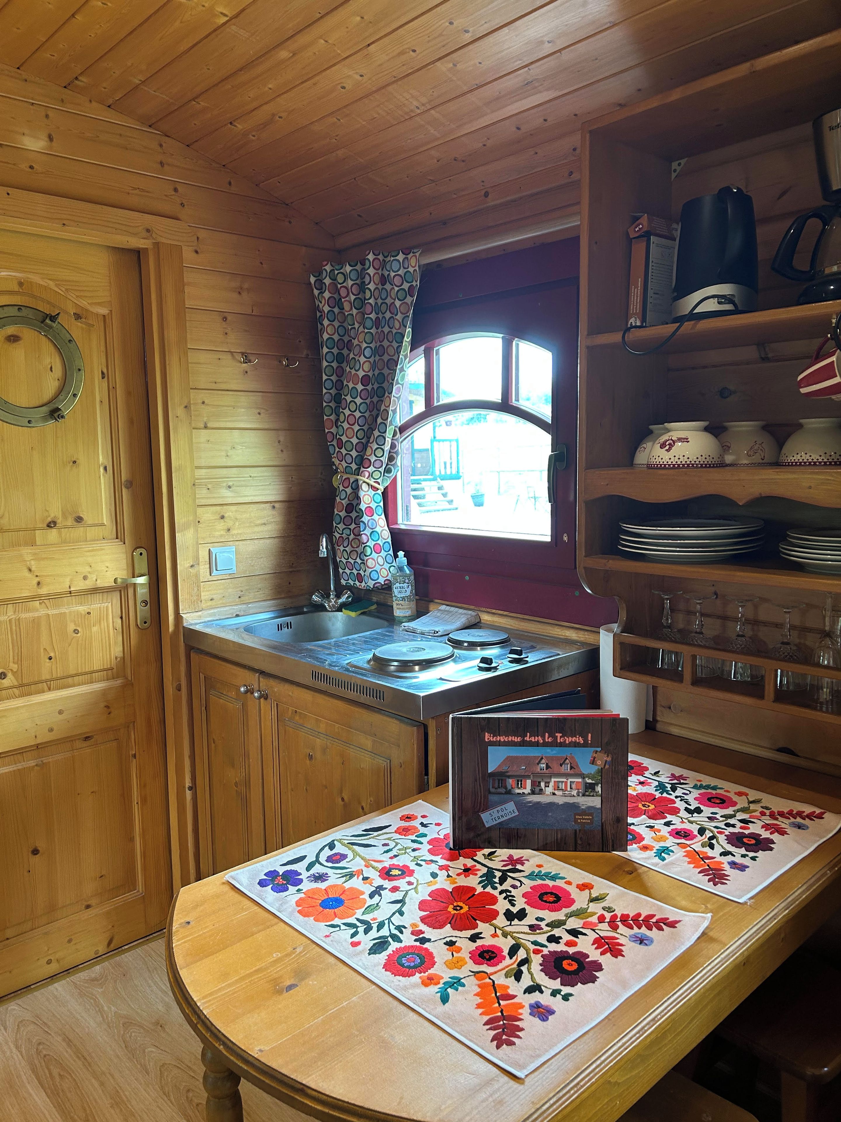 Interior em madeira com cozinha, janela com cortina e mesa com toalha de mesa florida.