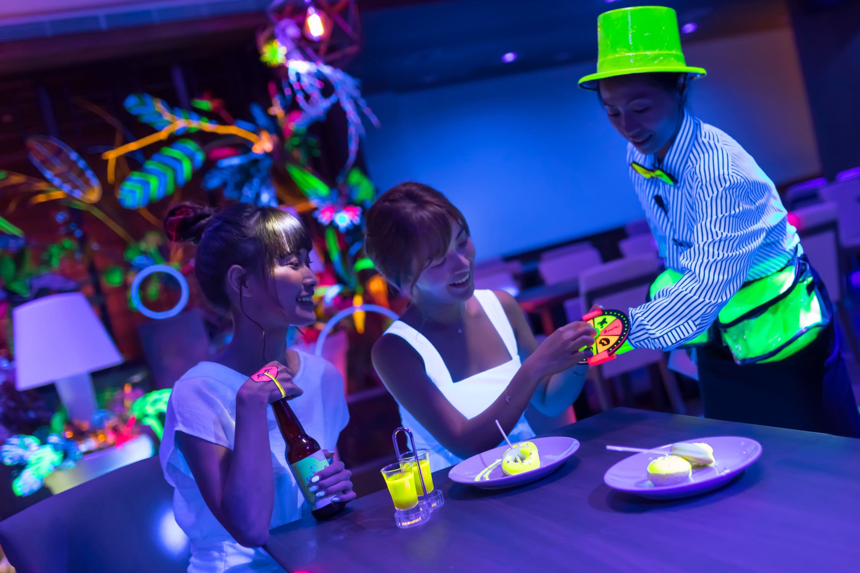 Um grupo de pessoas à mesa, com bebidas e pratos de comida, num espaço com decoração colorida e ambiente festivo.