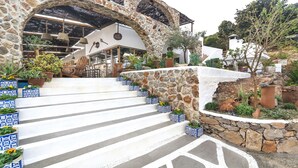 Uma escadaria de pedra leva a um edifício com interior rústico, rodeado por plantas em vasos e mesas ao ar livre.