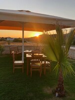 Um pátio ao ar livre com mesa e cadeiras, uma palmeira e um pôr do sol ao fundo.
