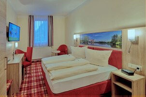 Quarto de hotel com cama, escrivaninha, cadeira, televisão e pintura de uma paisagem.