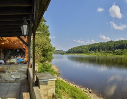 Restaurante à beira do rio com área externa, com vista para um rio calmo e colinas arborizadas.