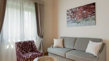 Uma sala de estar com um tapete estampado, um sofá cinza com almofadas brancas, uma poltrona estampada e uma mesa de centro de vidro.