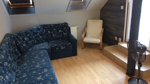 Sala de estar com sofá seccional azul, uma poltrona branca, luminária de chão preta e um ventilador preto. Há claraboias no teto e uma janela à direita.