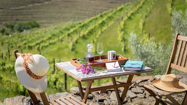 Uma mesa com um livro, óculos, uma garrafa e um tabuleiro em um terraço com vista para um vinhedo.