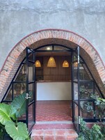 Uma entrada de tijolos em arco com portas de vidro que dá acesso a um espaço com armários de madeira e luminárias pendentes.