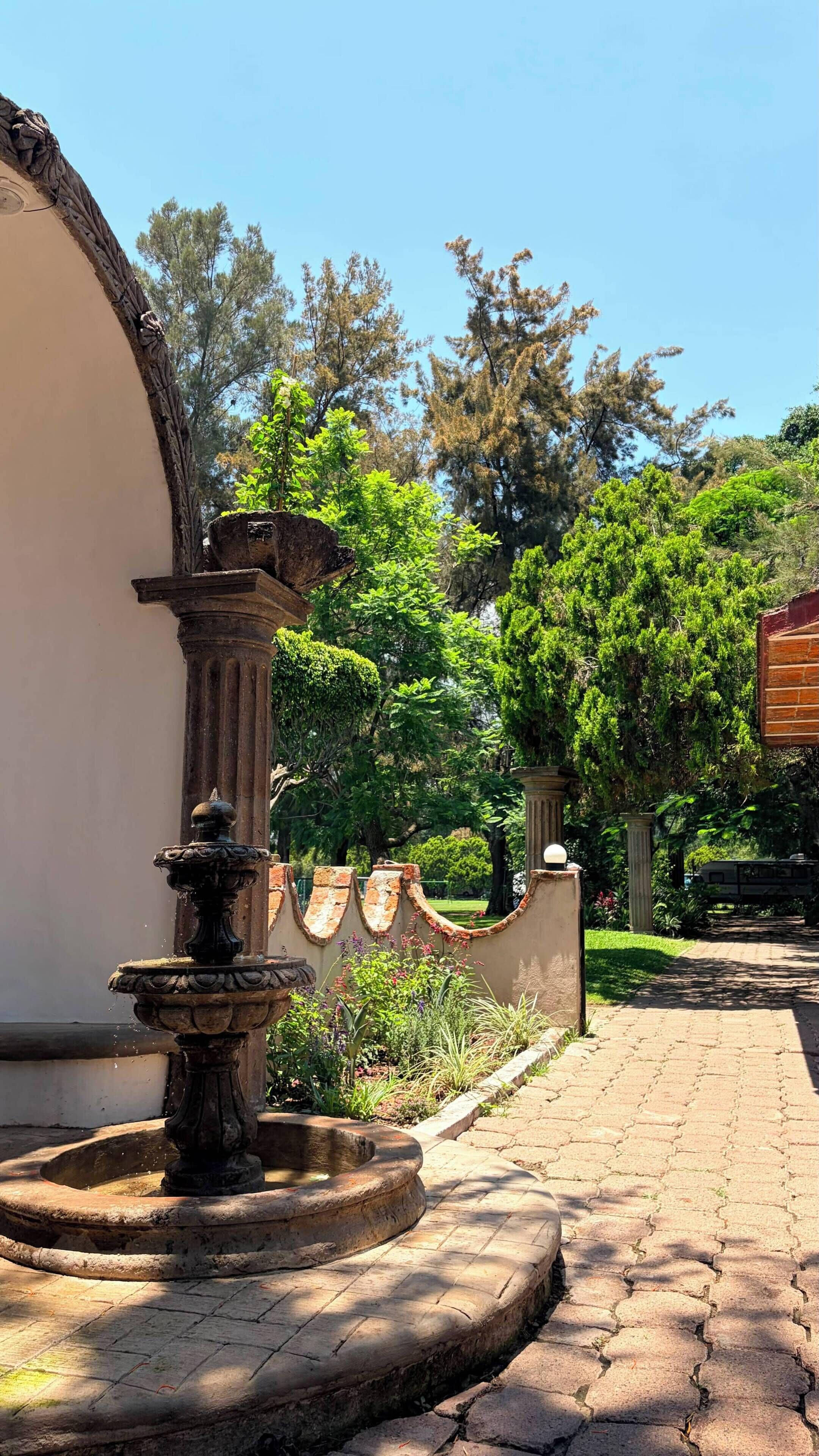 Um caminho no jardim com uma fonte, um muro de tijolos e vegetação exuberante.