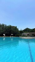 Uma piscina translúcida com espreguiçadeiras e guarda-sóis, cercada por vegetação exuberante e uma ponte de pedra.