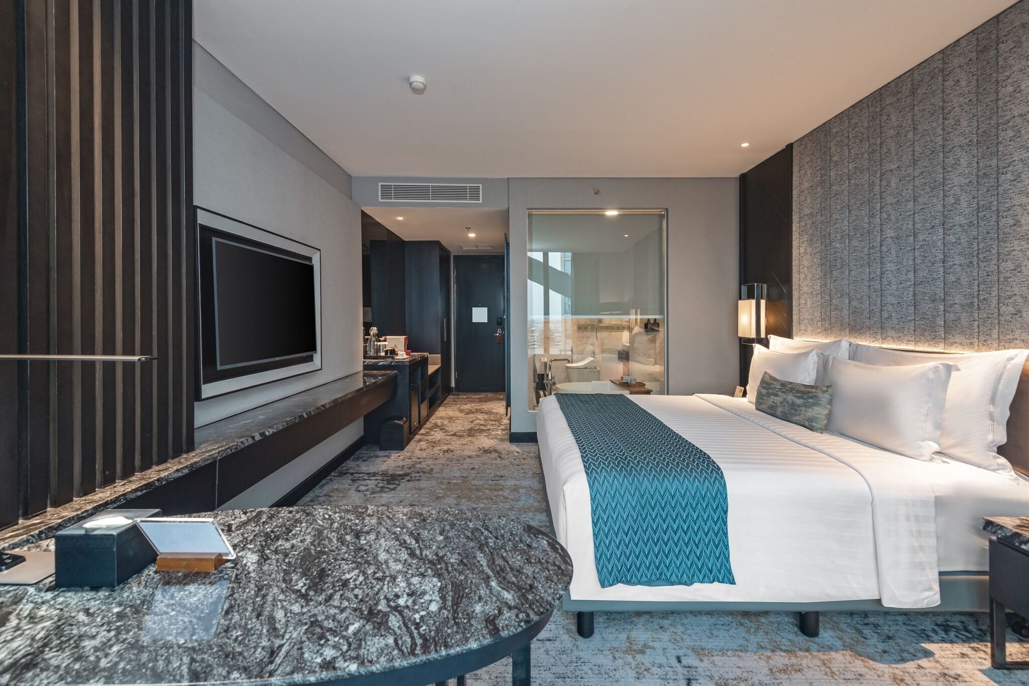 Quarto de hotel moderno com uma cama grande, televisão, área de estar e banheiro visível através de uma porta de vidro.