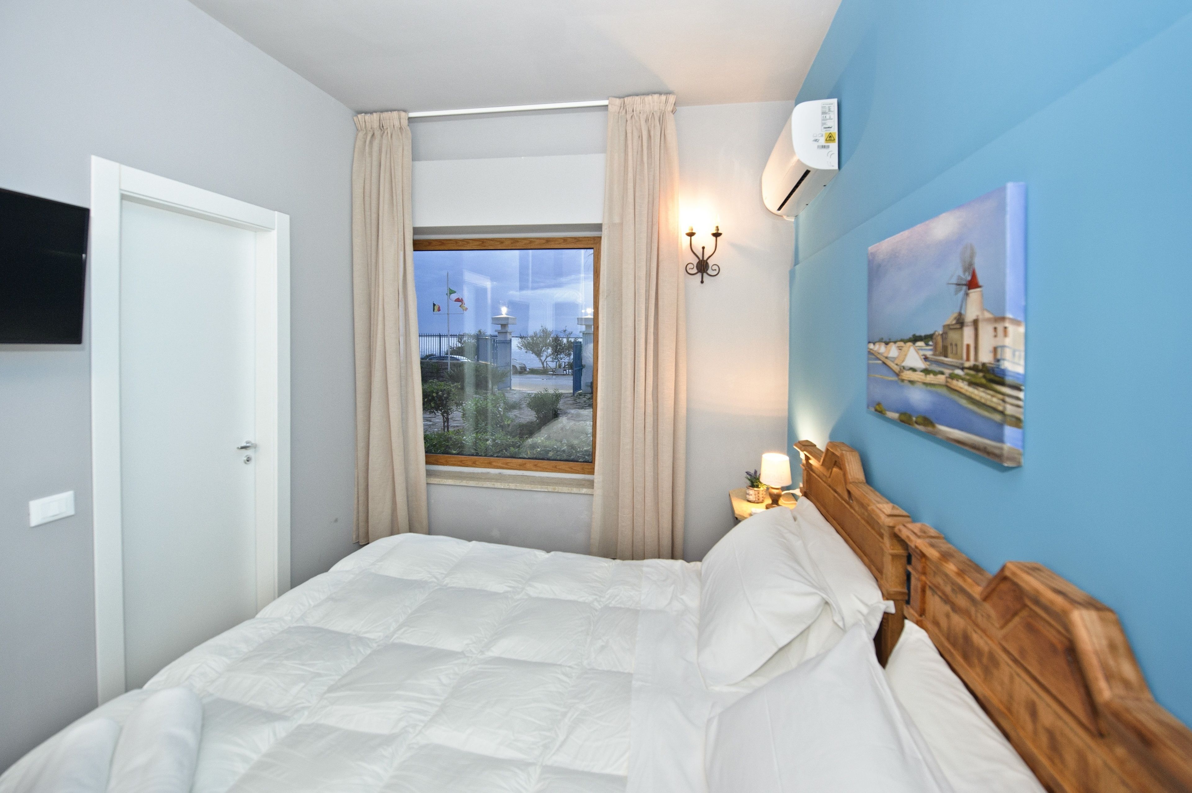 Um quarto com cama, janela com cortinas, ar-condicionado fixado na parede, mesa de cabeceira com abajur e um quadro na parede.