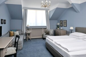 Quarto de hotel com uma cama grande, uma escrivaninha, uma cadeira, uma mesa pequena, um radiador e uma janela com cortinas.