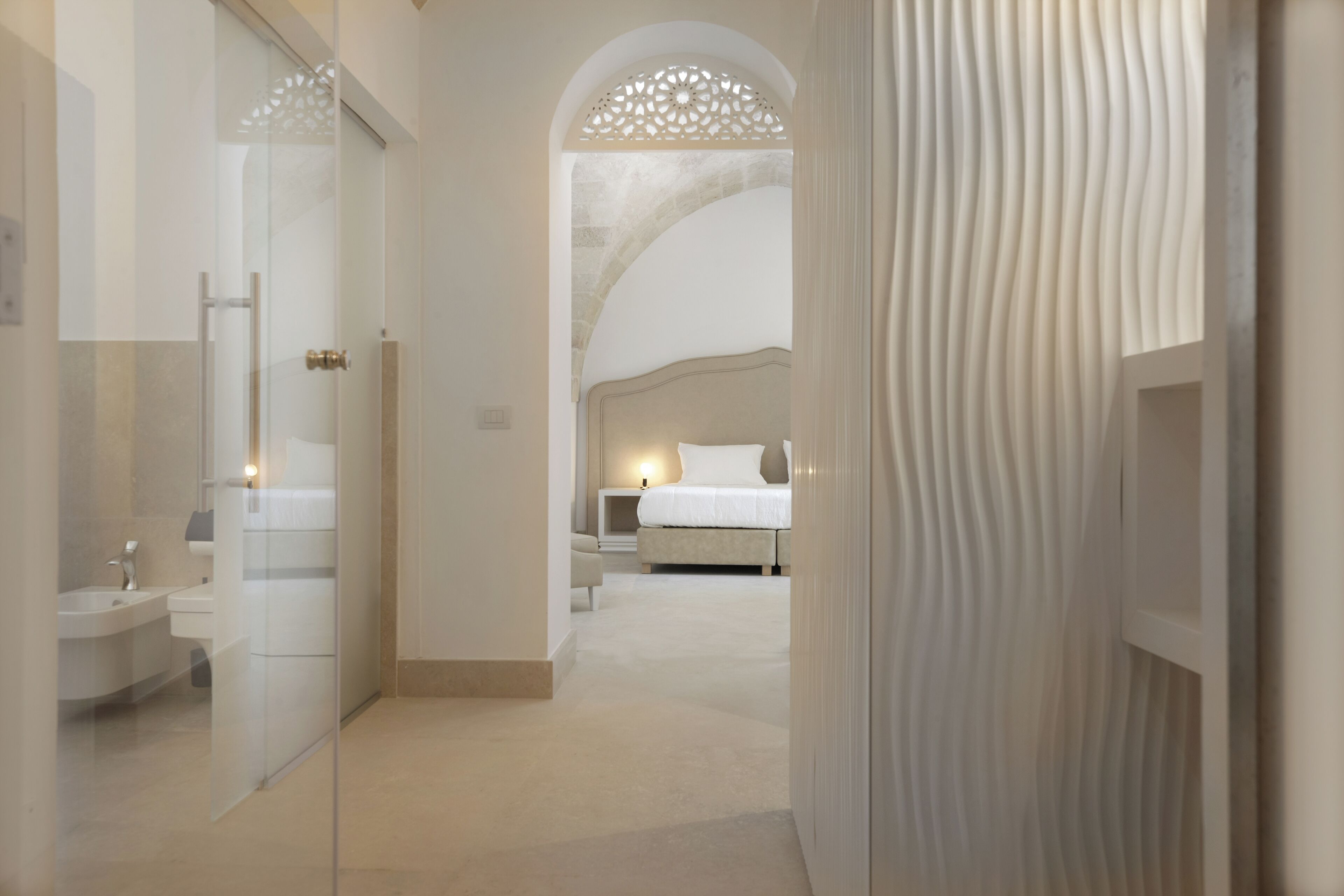Quarto de hotel moderno com banheiro, cama com cabeceira e vista por uma porta arqueada.