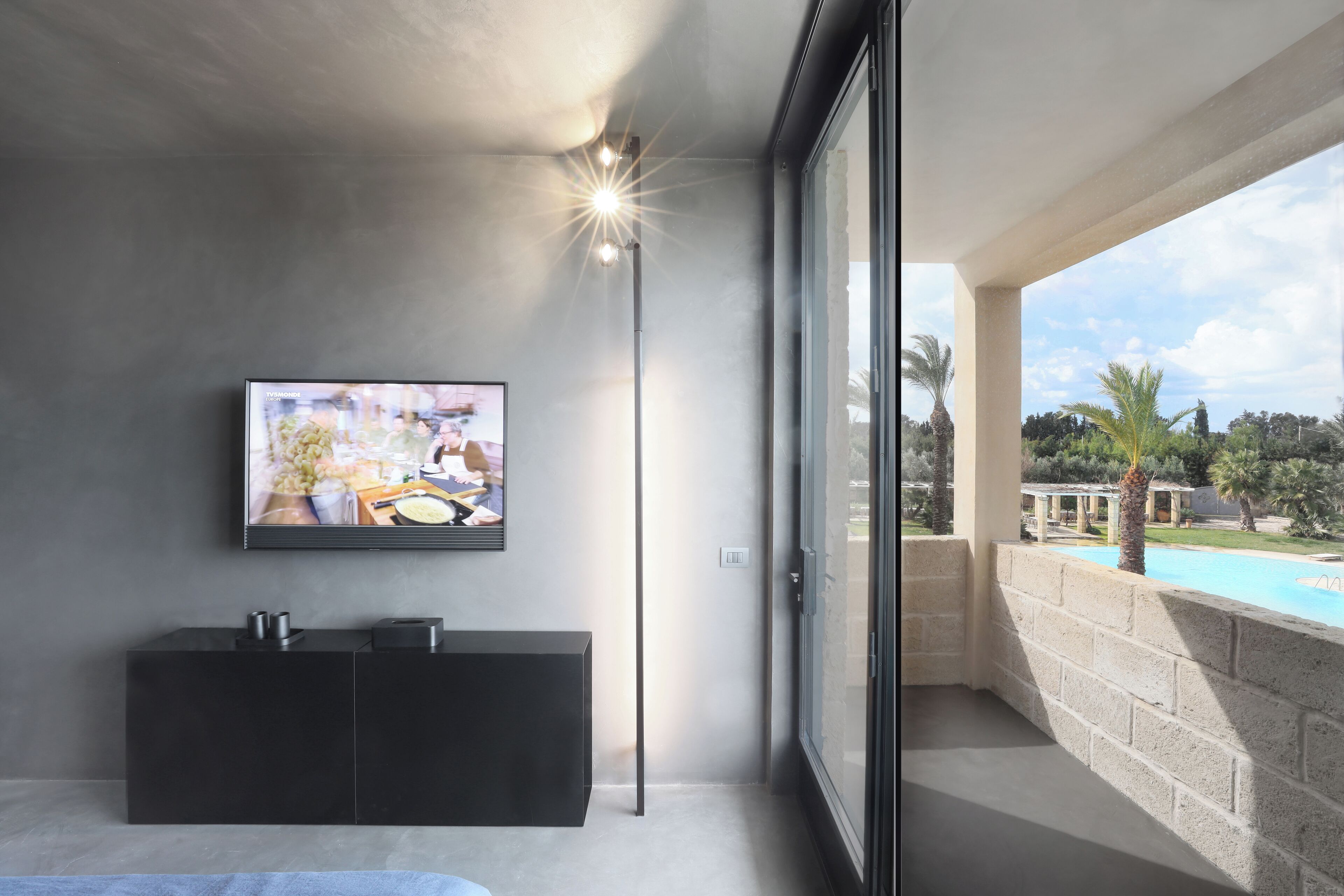 Uma sala de estar moderna com uma televisão de tela plana na parede, um rack de entretenimento preto e uma grande porta de vidro que dá para uma varanda com vista para a piscina.