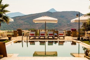 Piscina com cadeiras de descanso e guarda-sóis, cercada por muros de pedra e com vista para uma montanha.