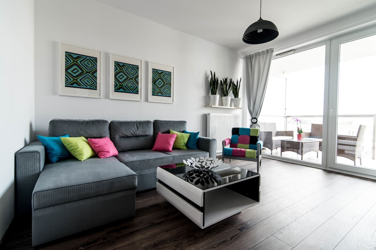 Uma sala de estar moderna com um sofá cinzento, almofadas coloridas, uma mesa de centro de vidro e janelas amplas com vista para o exterior.