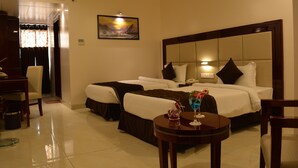 Quarto de hotel com uma cama grande, mesa de cabeceira com um copo de bebida, uma cadeira, escrivaninha e um quadro na parede.