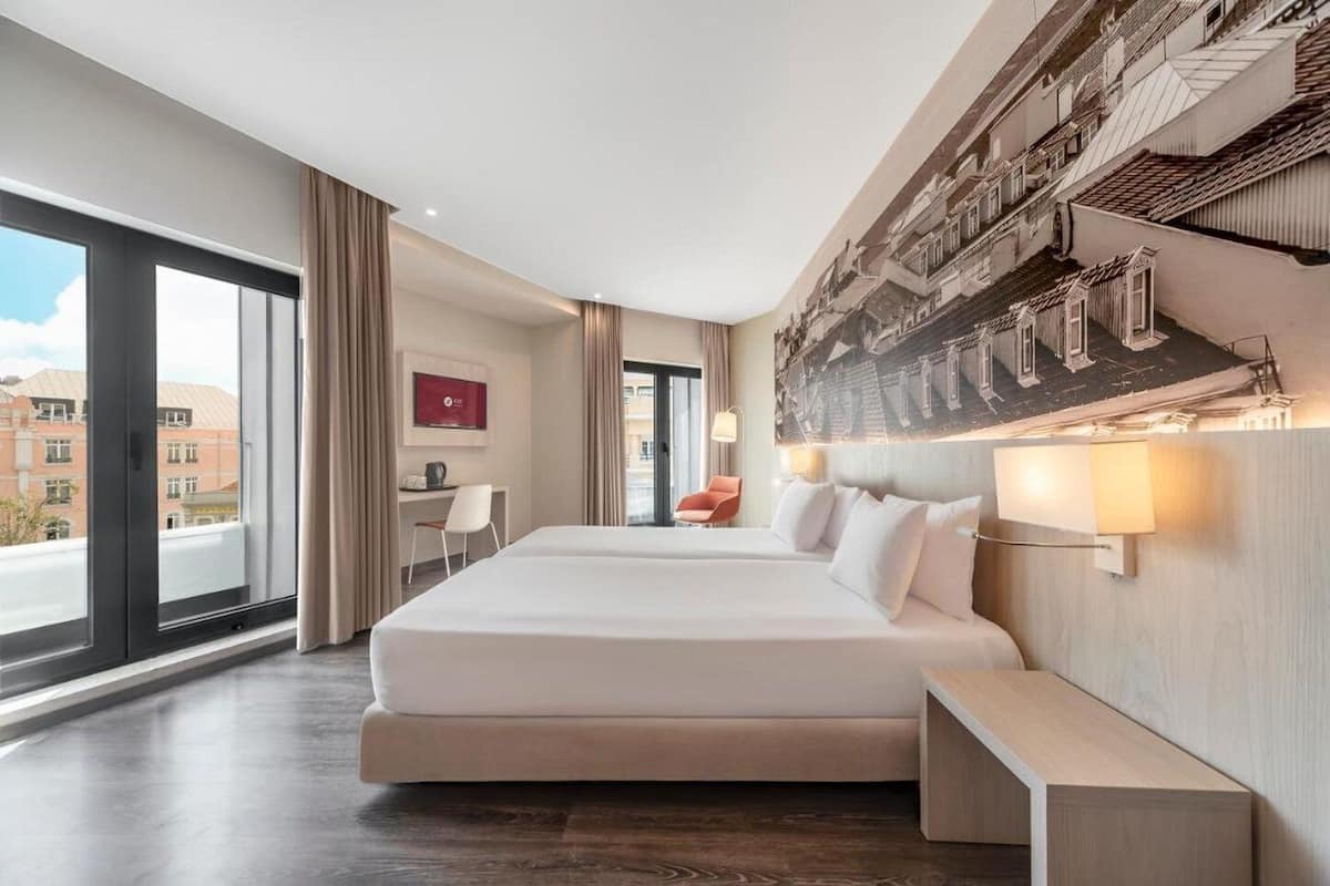 Quarto de hotel moderno com uma cama grande, uma escrivaninha e uma cadeira. Há uma janela ampla com vista para a cidade.