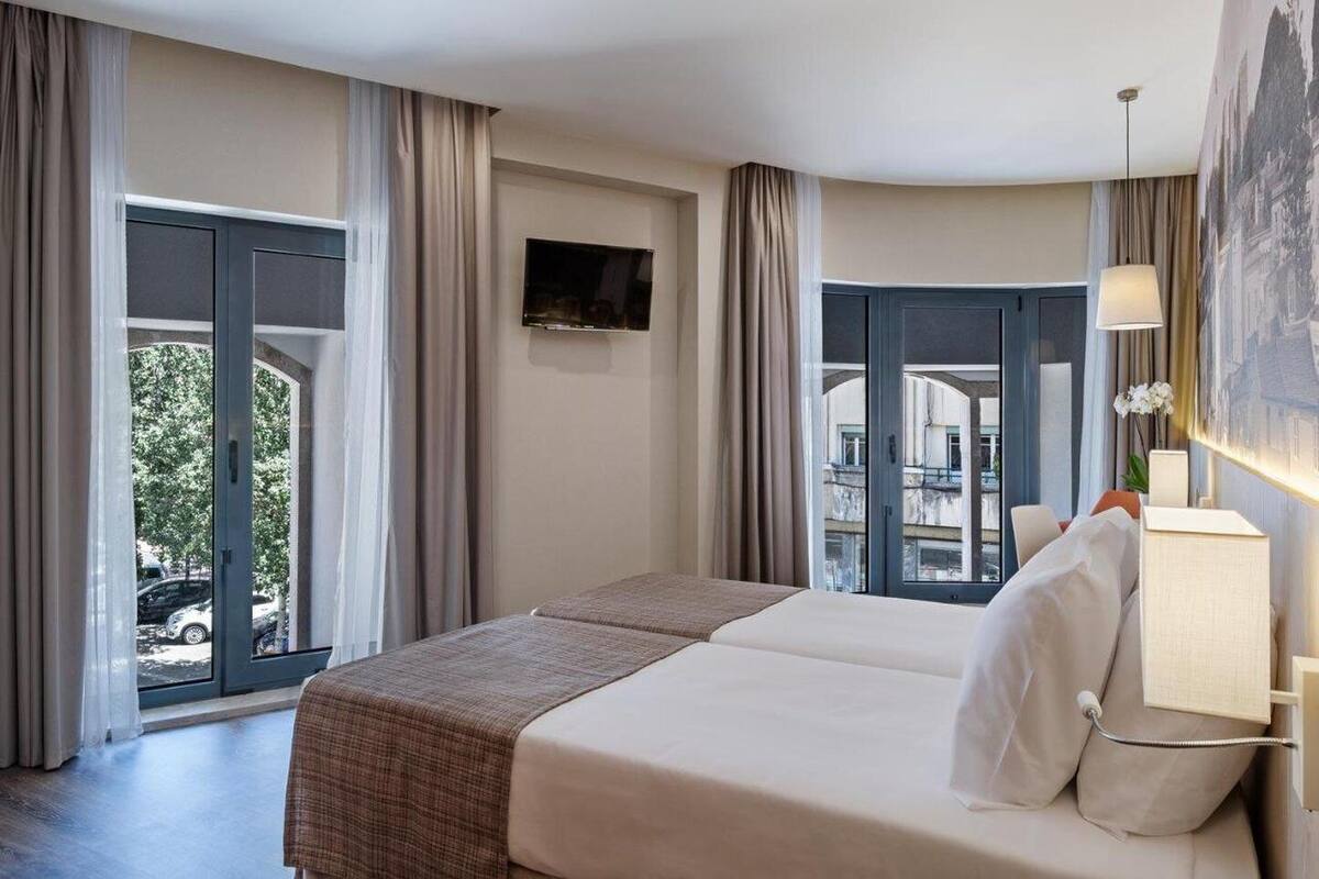 Quarto de hotel com uma cama grande, televisão na parede e vista para a cidade pelas janelas.