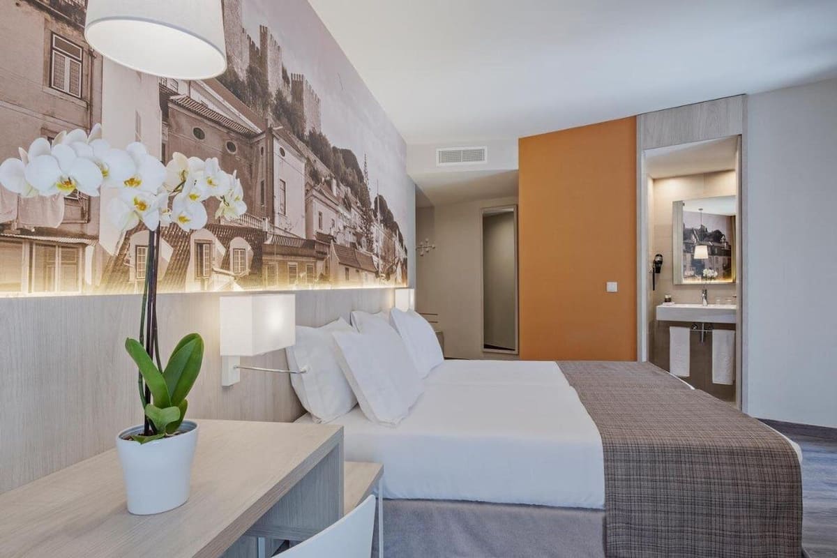 Quarto de hotel com cama, escrivaninha, orquídea em vaso, luminária na parede, banheiro com espelho e pintura de um edifício.