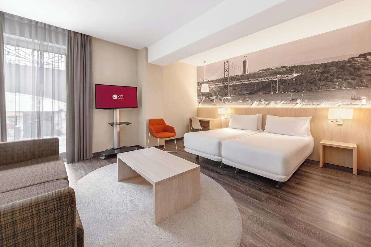 Quarto de hotel com cama, televisão, escrivaninha, cadeira e um quadro de uma ponte.