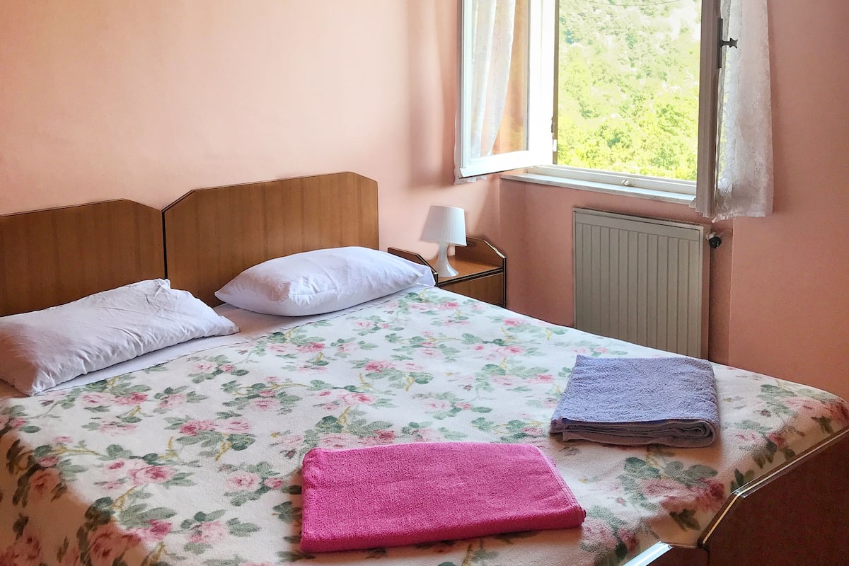 Quarto com cama, mesa de cabeceira, abajur e janela com cortinas.