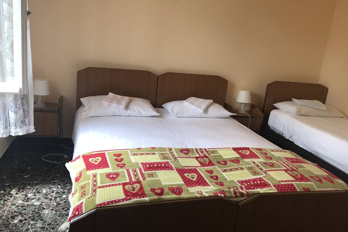 Quarto de hotel com duas camas de solteiro, uma cama grande com colcha, mesa de cabeceira com abajur e janela com cortina.