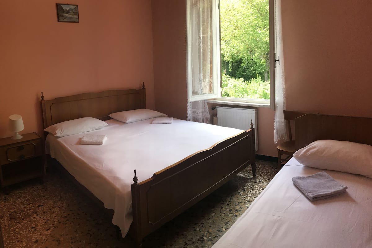 Quarto com cama de madeira, mesa de cabeceira, janela com vista para o verde e radiador.