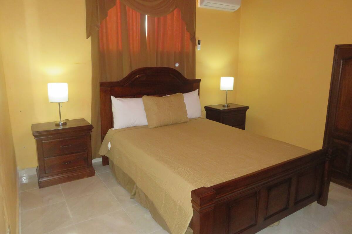 Um quarto com cama de madeira, duas mesas de cabeceira com luminárias, um criado-mudo, uma janela com cortinas e um aparelho de ar condicionado.