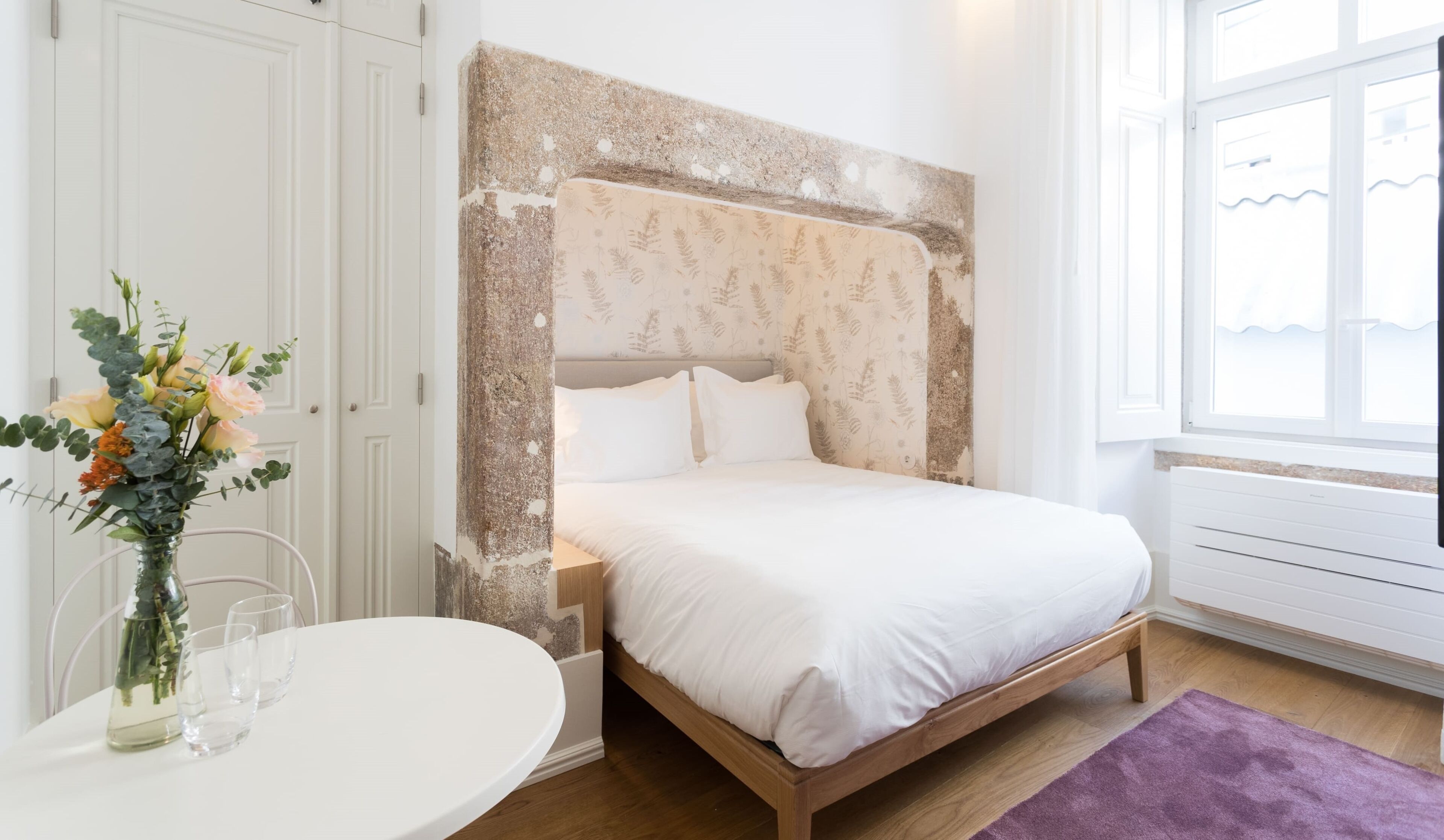 Quarto com cama de madeira, roupa de cama branca, uma mesa pequena com um vaso de flores e uma janela com cortinas.