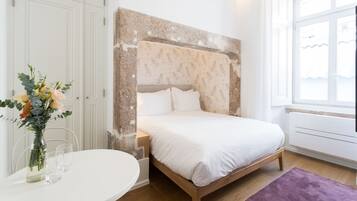 Quarto com cama de madeira, roupa de cama branca, uma mesa pequena com um vaso de flores e uma janela com cortinas.