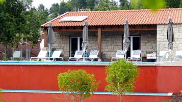 Uma área ao lado da piscina com espreguiçadeiras e guarda-sóis, um telhado de azulejos e uma parede de pedra.