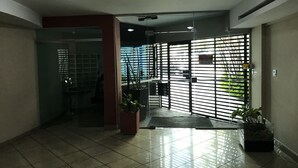 Um saguão com piso de azulejos, porta de vidro com portão de segurança metálico e plantas em vasos.