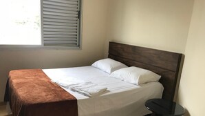 Uma cama bem arrumada com roupa de cama branca e um cobertor marrom, uma pequena mesa de cabeceira e uma janela com persianas.